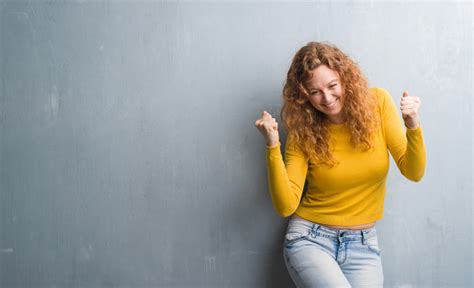 젊은 빨강 머리 여자 매우 행복 하 고 흥분 무기와 우승자 제스처를 하 고 회색 그런 지 벽에 웃 고 성공을 위한 비명 축 하 개념입니다 곱슬 머리에 대한 스톡 사진 및