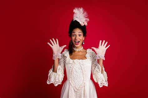 우아한 바로크 양식의 가운을 입은 즐거운 여성이 생생한 붉은 배경을 배경으로 장난기 넘치는 몸짓으로 놀라움을 선사합니다 25 29세에 대한 스톡 사진 및 기타 이미지