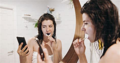 소셜 미디어가 영향을 미칩니다 하얀 수건을 쓴 젊은 행복한 여자가 이를 닦고 둥근 거울에 있는 세련된 욕실에서 전화 화면을