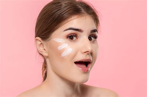 아름다운 모델은 분홍색 배경 위에 고립 된 히알루론산 크림의 흰색 튜브를 들고 피부에 보습 화장품을 적용합니다 미용 및 건강 치료의 개념 갈색 머리에 대한 스톡 사진 및