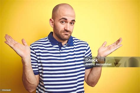 노란색 고립 된 배경 단서와 팔과 손을 제기 혼란 표정 위에 캐주얼 스트라이프 블루 티셔츠를 입고 수염을 가진 젊은 대머리 남자 의심 개념 T 셔츠에 대한 스톡 사진 및