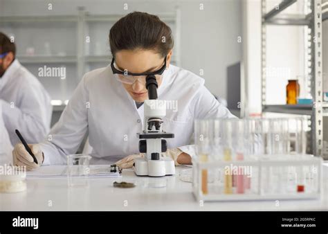 Scientist Looking In Microscope And Taking Notes While Working In Microbiology Laboratory Stock