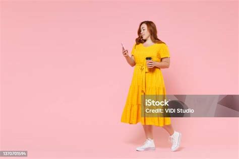 전체 길이 의 초상화 매력적인 젊은 빨간 머리 플러스 크기 몸 긍정적 인 여성 20 S 노란색 드레스 홀드 종이 컵 커피 또는 차 파스텔 핑크 컬러 배경 스튜디오에서 격리