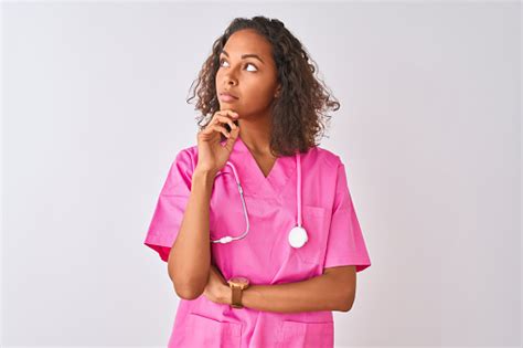 청진기를 입은 젊은 브라질 간호사 여성이 고립된 흰색 배경 위에 서서 턱에 손을 얹고 질문 펜심 표현을 생각하고 있습니다 사려 깊은 얼굴로 미소 의심 개념 간호사에 대한