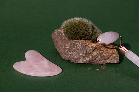 เครื่องนวดลูกกลิ้งหน้าควอตซ์และมีดโกน Gua Sha บนหินที่มีตะไคร่น้ําเติบโตพื้นหลังสีเขียวแยกไ ภาพ