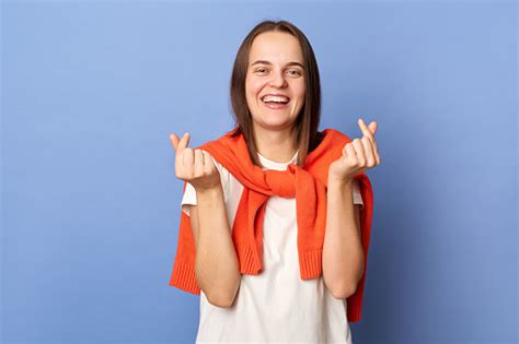 흰색 티셔츠를 입고 파란색 배경에 고립된 어깨 위에 스웨터를 묶은 매우 행복한 낭만적인 여성의 실내 촬영 미니 사랑의 제스처를 보여주며 긍정적인 감정을 표현합니다 30 34