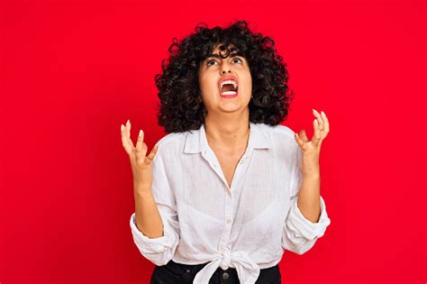 고립 된 빨간 배경 에 흰색 캐주얼 셔츠를 입고 곱슬 머리를 가진 젊은 아랍 여성은 미친 미친 미친 소리와 공격적인 표정과 팔을 제기 고함 좌절 개념입니다 감정에 대한 스톡