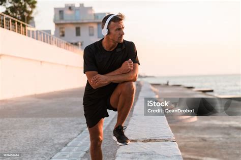 바닷가 근처를 걷고 일출 중에 무선 헤드폰을 통해 음악을 듣고 반바지와 티셔츠를 입은 근육질의 스포티한 남자 30대 의 이미지 30 39세에 대한 스톡 사진 및 기타 이미지