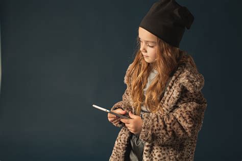 진한 파란색 배경의 스튜디오에서 따뜻한 겨울 옷을 손에 들고 태블릿을 손에 든 빨간 머리 소녀 모피 코트와 모자에 긴 머리카락을 가진 어린 소녀 초상 5g에 대한 스톡 사진