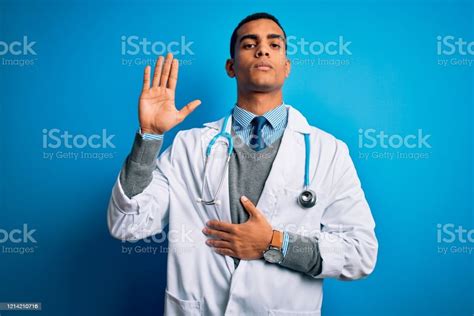 가슴과 열린 손바닥에 손으로 맹세 충성 약속 선서를 만들기 파란색 배경 에 코트와 청진기를 입고 잘 생긴 아프리카 계 미국인 의사 남자 의사에 대한 스톡 사진 및 기타