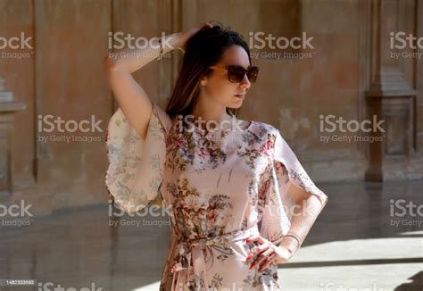 꽃이 만발한 밝은 분홍색 여름 드레스와 선글라스를 쓴 젊은 갈색 머리 여성의 초상화는 돌 배경으로 야외에 서서 포즈를 취하고 있다 스페인 유럽 1 여자만에 대한 스톡 사진