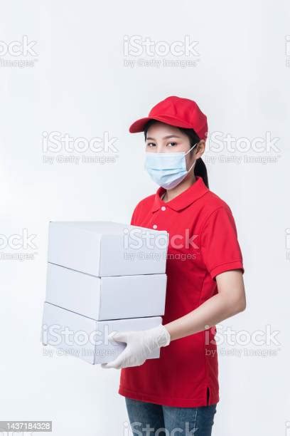 빨간 모자 빈 티셔츠 유니폼 얼굴 마스크 장갑을 낀 의식이 있는 젊은 배달원의 이미지는 밝은 회색 배경 스튜디오에 격리된 빈 흰색 판지 상자 팬데믹 코로나바이러스 바이러스