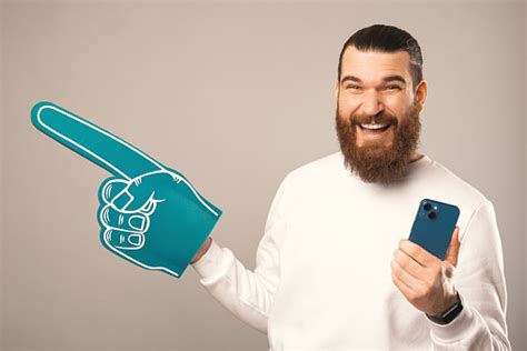 쾌활한 수염을 가진 남자가 파란색 팬 폼 장갑을 착용하고 전화를 들고 있습니다 갈색 머리에 대한 스톡 사진 및 기타 이미지