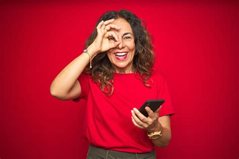 빨간 고립 된 배경을 통해 스마트 폰을 사용하는 중년 노인 여성 행복한 얼굴은 손가락을 통해 보고 눈을 통해 손에 확인 기호를하고 미소 보여주기에 대한 스톡 사진 및 기타