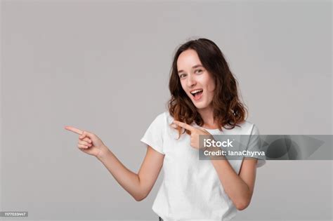 회색 배경 위에 고립 된 프레임의 왼쪽에있는 빈 공간을 가리키는 흰색 티에 예쁜 갈색 머리 여자 광고 의 공간을 복사합니다 가리키기에 대한 스톡 사진 및 기타 이미지
