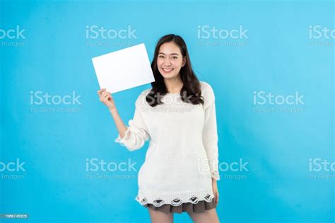 긴 머리를 가진 아시아 여성은 눈을 마주치고 흰 종이 표지판을 들고 얼굴을 닫고 미소를 지으며 코트와 치마를 입는다 스튜디오에서 파란색 장면을 사진으로 찍습니다 25 29