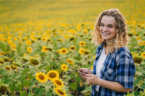 해바라기 밭 앞에 서서 태블릿으로 작업하는 카메라를 보며 웃고 있는 매력적인 곱슬머리 여자가 피는 해바라기를 검사합니다 공간을 복사합니다 농업 노동자 현대 기술 농업