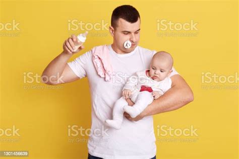 어린 아버지는 아기를 안고 우유로 아이를 먹이려고 하고 아이의 젖꼭지를 손에 들고 포즈를 취하고 아무렇지 않게 입고 있는 남자가 노란 배경위에 고립된 수유병 아빠 아기를