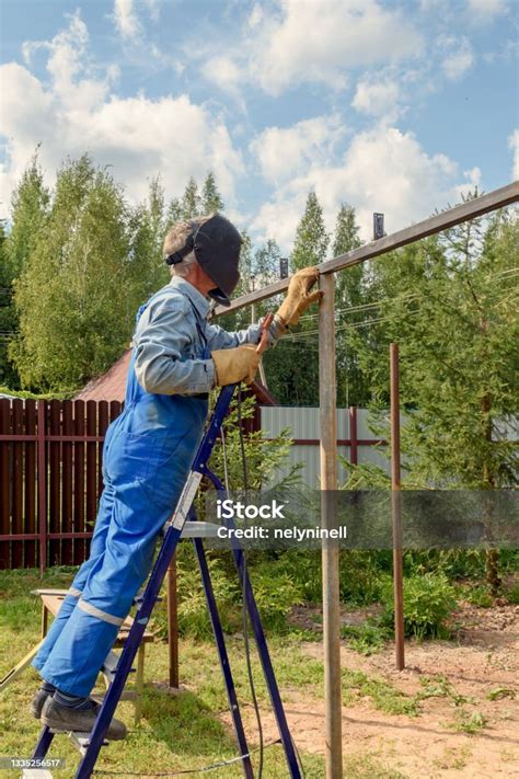 용접 마스크 건설 유니폼 및 보호 장갑을 착용한 남자 용접기는 거리 건설 현장에서 금속을 요리합니다 여름날 시골 집 근처 파빌리온 퍼골라 건설 강철에 대한 스톡 사진 및