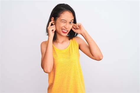 노란 캐주얼 티셔츠를 입은 젊은 중국 여성이 시끄러운 음악 소리에 짜증나는 표정으로 손가락으로 귀를 덮는 고립된 흰색 배경 위에 서 있습니다 청각 장애인 개념입니다 T