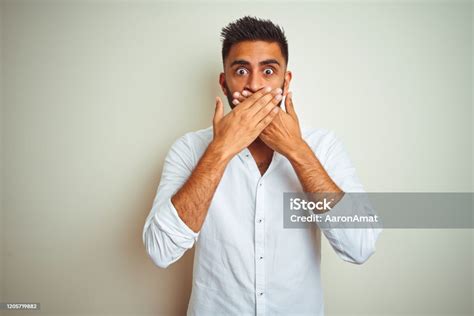 고립 된 흰색 배경 위에 서있는 우아한 셔츠를 입은 젊은 인도 남자는 실수로 손으로 입을 덮는 충격을 받았습니다 비밀 개념입니다