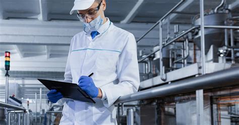 모자 마스크 및 흰색 코트의 기술자가 생산 공장에 서서 시트에 펜으로 메모를합니다 검사관은 낙농 공장에서 표시기를 제거합니다 엔지니어는 생산에 대한 통계를 유지합니다