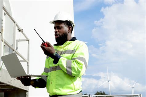 안전 조끼와 헬멧을 쓴 아프리카 엔지니어는 워키 토키를 사용하고 풍력 터빈 농장에서 노트북 컴퓨터에서 작업합니다 작업자가 구름 낀 하늘 풍차 공원에서 일하고 있습니다