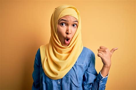 노란 배경 위에 아랍 전통 히잡을 입고 곱슬 머리를 가진 젊은 아름다운 여자는 측면에 손 손가락으로 가리키는 놀라움 입을 벌리고 표정 가리키기에 대한 스톡 사진 및 기타