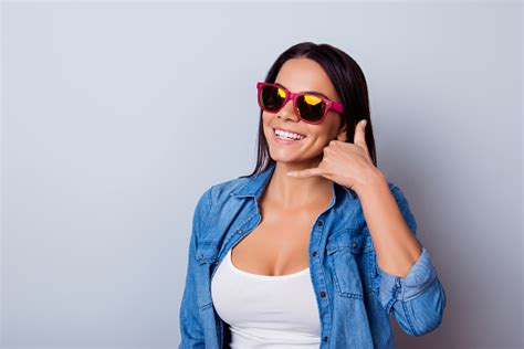전송 해 미소 밝은 라틴 여자는 손으로 그녀의 전화를 몸짓 이다 그녀는 캐주얼 청바지 셔츠와 흰색 내의 선글라스 착용 하 고 밝은 배경에 스탠드 갈색 머리에 대한 스톡 사진