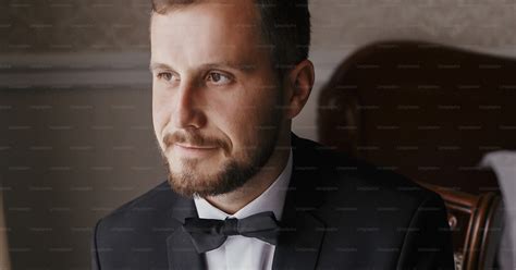 검은 양복과 나비 넥타이를 맨 세련된 신랑이 창문 조명에 의자에 앉아 있습니다 남자의 자신감 있고 행복한 초상화 아침에