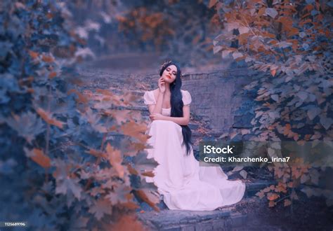 긴 하얀 드레스를 입고 그녀의 머리에 우아한 황금 화 환 그녀의 눈을 폐쇄 검은 머리 여자와 꿈 돌 계단에 숲 여신 동화 정신을 잔디의 소리를 수신 대기 하 고 나뭇잎