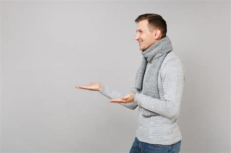 웃 고 회색 스웨터 스카프 가리키는 손을 옆으로 회색 벽 배경에 고립에서 젊은 남자의 측면 보기 건강 패션 라이프 스타일 사람들 진심으 감정 추운 계절 개념 복사 공간을