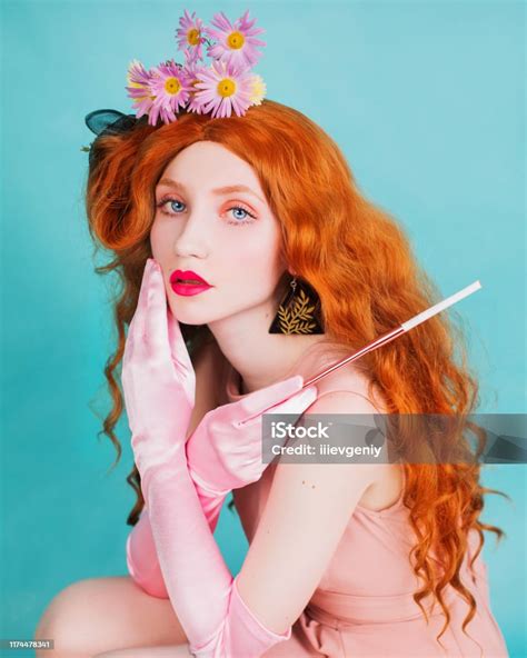 빨간 곱슬 머리에 빨간 입술과 꽃 르네상스 빨간 머리 모델 아가씨는 마우스 피스를 손에 쥐고 있습니다 파란색 배경에 스튜디오에서 긴 머리와 분홍색 장갑아름다운 빨간 머리