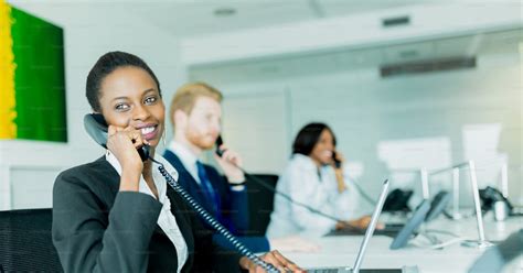 사무실의 콜센터에서 일하는 아름답고 흑인이며 젊은 여성이 책상 반대편에 빨간 머리 파트너와 함께 다른 고객과 이야기하고 있습니다