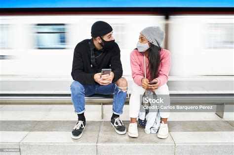 가면과 양모 모자를 쓴 젊은 인종 간 부부가 지하철 플랫폼에 앉아 스마트폰을 손에 들고 있다 마차 기차 지하철 운동 관광객에