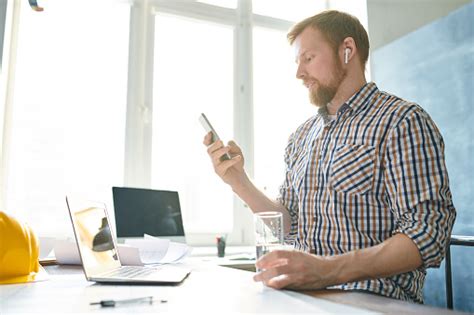 휴대 전화에서 메시지를 확인 하는 동안 테이블에 앉아서 유리에서 물을 마시는 심각한 바쁜 젊은 수염 엔지니어 무선 이어버드 건설 산업에 대한 스톡 사진 및 기타 이미지
