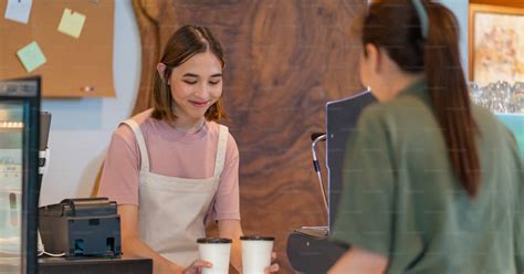 젊은 아름 다운 아시아 여자 커피 숍 직원 바리 스타가 카페에서 일하고 있습니다 웃는 여성 웨이트리스는 고객에게 종이 커피 컵에 테이크 아웃 주문을 준비합니다 중소 기업
