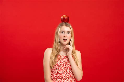 열린 눈을 가진 십대 소녀는 머리에 누워있는 사과 빈 빨간색 배경 무료 복사 공간으로 서 있습니다 깜짝 놀랄만한 감정으로 꽃무늬 프린트 드레스를 입은 영리한 아이의 초상화