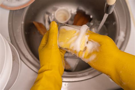 노란색 보호 고무 장갑에 손을 닫고 거품과 식기로 가득 찬 부엌 싱크대에 대해 흐르는 수돗물 아래에서 마시는 유리를 씻습니다 개수대에 대한 스톡 사진 및 기타 이미지