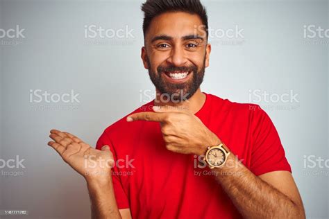 고립 된 흰색 배경 위에 빨간 티셔츠를 입은 젊은 인도 남자는 손으로 손가락으로 가리키는 동안 카메라에 놀랍고 미소를 지었습니다