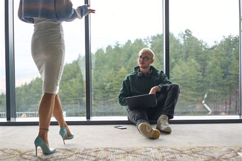 현대식 사무실 바닥에 아무렇지도 않게 앉아서 창문에 기대어 노트북을 사용하고 있는 금발 남자와 매력적인 여성 동료가 근처에 서 있습니다 2명에 대한 스톡 사진 및 기타