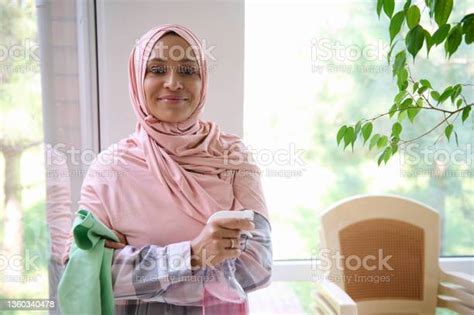 아름다운 아랍 무슬림 여성이 집의 봄 청소 중에 창문을 씻기위한 세제 스프레이와 손에 걸레를 들고 히잡을 쓴 머리를 가진 아름다운 아랍 무슬림 여성 카메라를보고 미소 가사