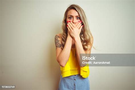 흰색 고립 된 배경 위에 서있는 노란색 티셔츠를 입은 젊은 아름다운 여성은 실수로 손으로 입을 덮는 충격을 받았습니다 비밀 개념입니다 T 셔츠에 대한 스톡 사진 및 기타