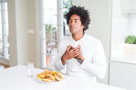 아프리카계 미국인 배고픈 남자 점심 식사를 위해 햄버거를 먹고 얼굴에 닫힌 눈과 감사 제스처와 가슴에 손으로 웃 고 건강