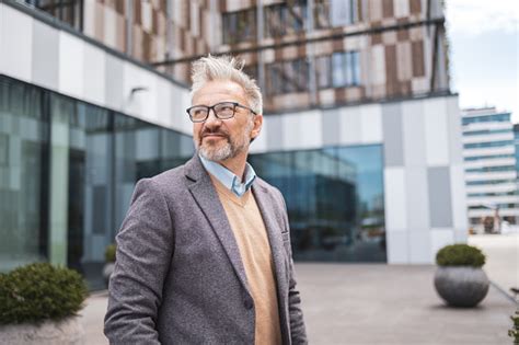 안경과 턱수염을 기른 도시 환경의 성숙한 사업가 블레이저와 스웨터를 입은 그는 자신감과 프로페셔널리즘을 발산한다 배경에 현대 건물이 있는 도시 풍경입니다 비즈니스에 대한