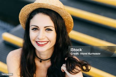 어두운 고급스러운 머리 매력적인 밝은 눈 잘 생긴 빨간 입술과 순수한 피부가 손으로 그녀의 머리를 만지고 여름 모자를 쓰고 야외에서 카메라에 즐겁게 미소 짓는 행복한 여성