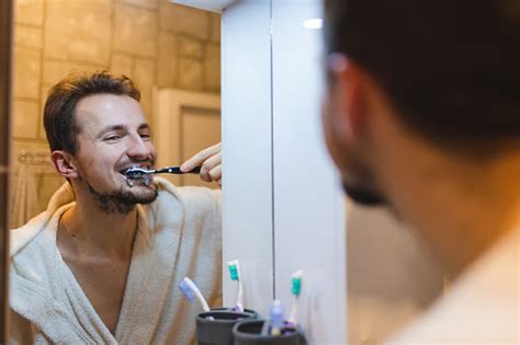 화장실 거울에 자신을 보면서 그의 이빨을 닦는 목욕 가운을 입은 젊은 남자 25 29세에 대한 스톡 사진 및 기타 이미지 25 29세 Brand Name Smart