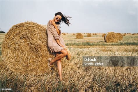 건초 베일 근처의 밀 밭에서 포즈를 취하는 선글라스와 밀짚 모자를 쓴 긴 머리를 가진 아름다운 소녀 여름 드레스에 행복한 갈색 머리 9월에 대한 스톡 사진 및 기타 이미지