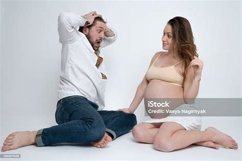 흰색 셔츠를 입은 충격을 받은 청년이 그의 머리에 손을 얹고 하얀 배경 위에 고립된 아름다운 임신한 아내를 바라보고 있다 Health에 대한 스톡 사진 및 기타 이미지