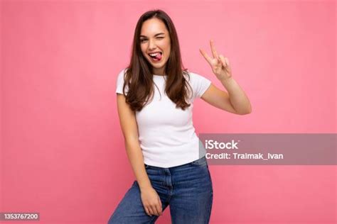 분홍색 배경에 고립 된 모형을위해 캐주얼 한 흰색 티셔츠를 입고 두 손가락을 보여주는 진지한 감정을 가진 젊은 행복한 긍정적 인 아름다운 갈색 머리 여성의 초상화 사진 T